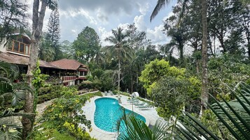 Piscina rodeada por vegetação exuberante e um edifício de dois andares com telhado em telhas.