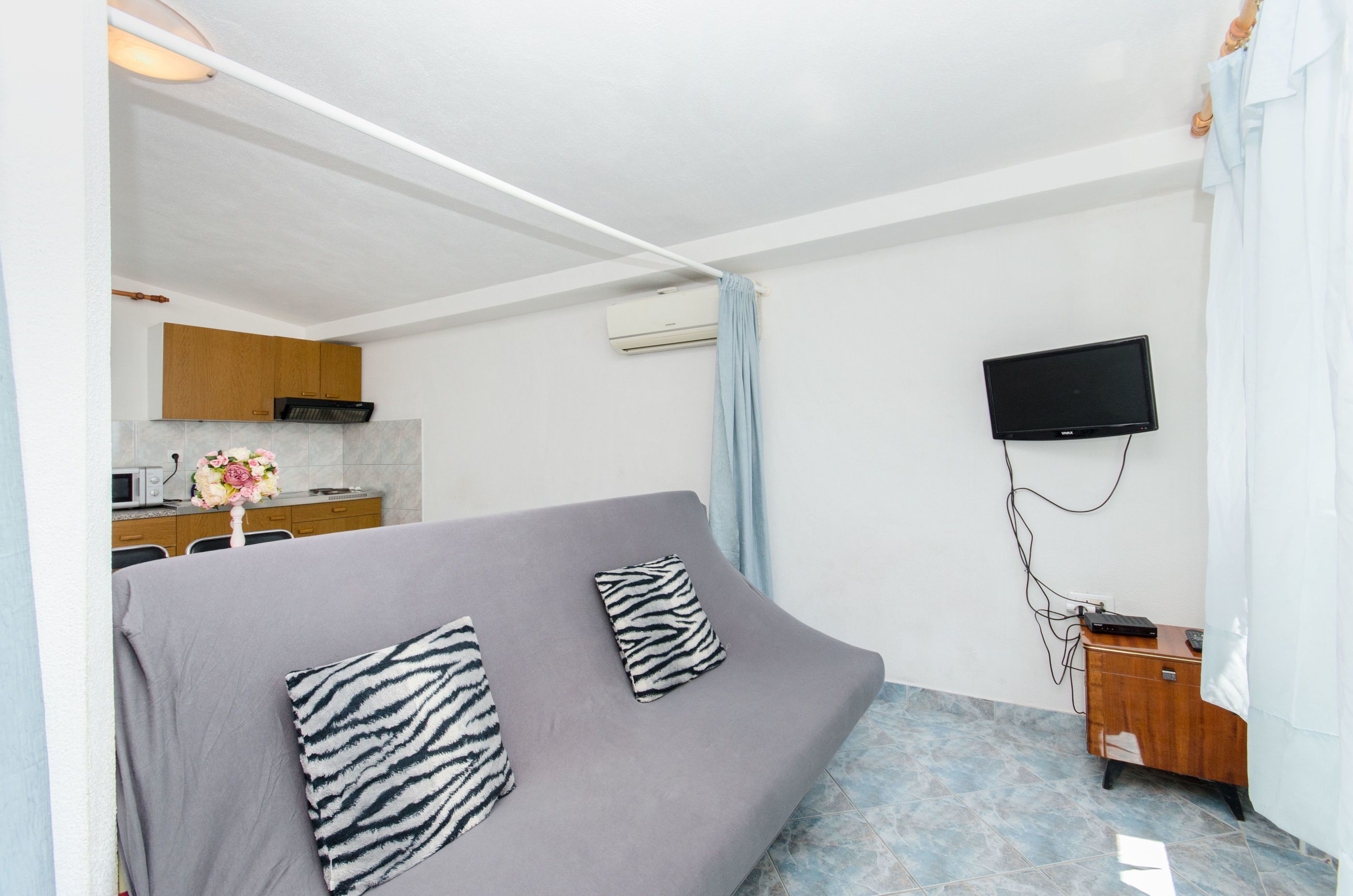 Quarto moderno com um sofá cinzento, duas almofadas com padrão de zebra, um armário de madeira, uma TV de tela plana fixada na parede e uma pequena mesa lateral de madeira.