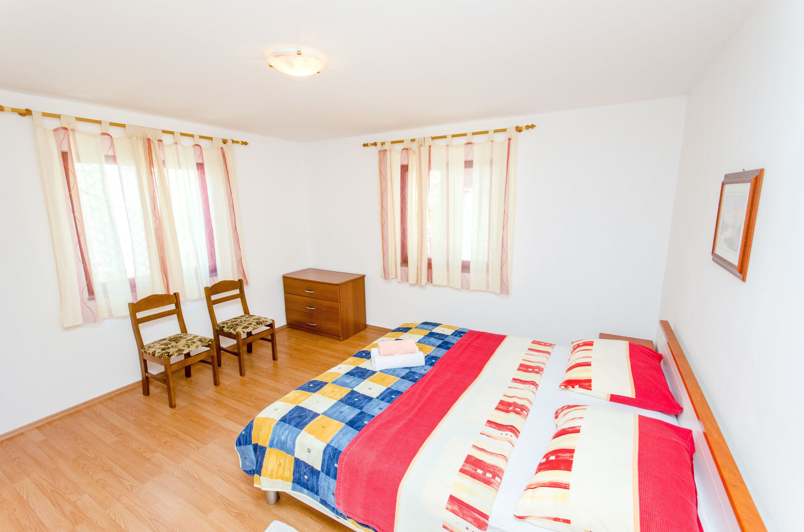 Quarto com cama, duas cadeiras, mesa de cabeceira e janela com cortinas.