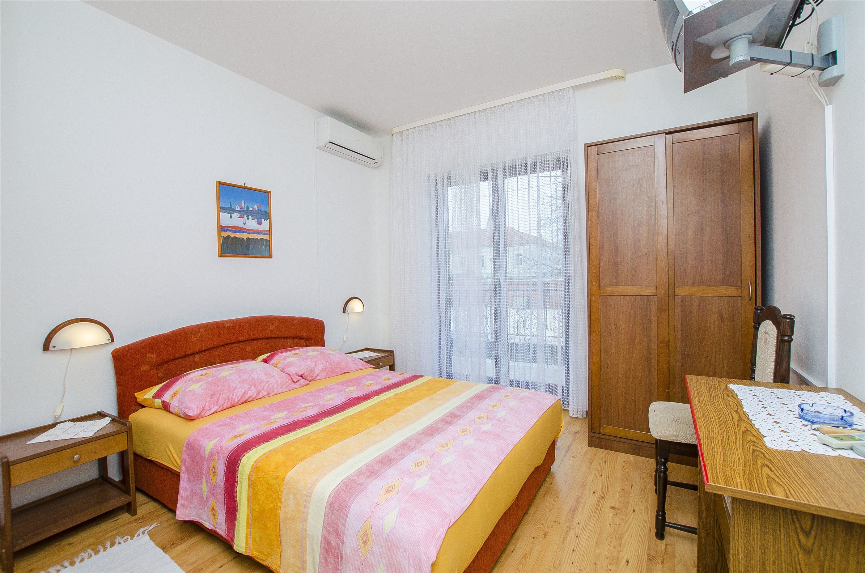 Quarto com cama, mesas de cabeceira, guarda-roupa de madeira, escrivaninha com cadeira e janela com cortinas.