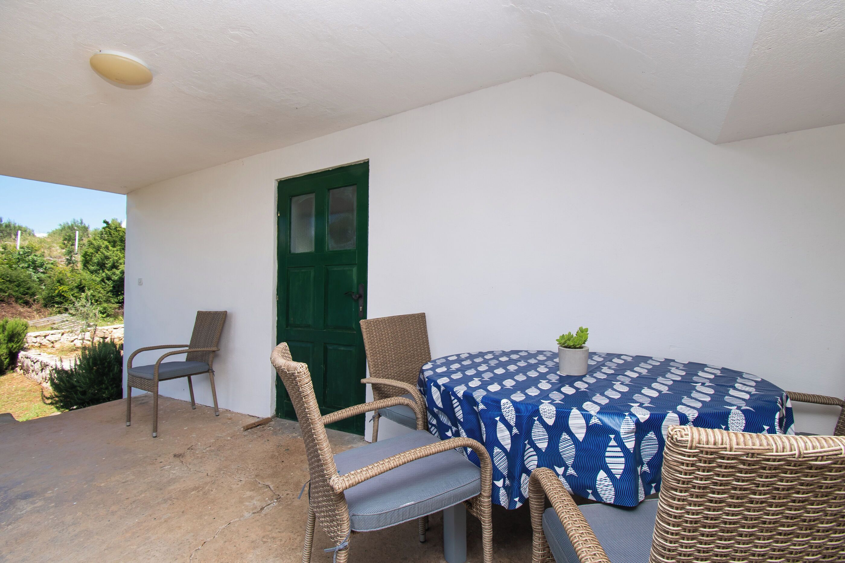 Um pátio com móveis de vime, uma mesa com toalha de mesa estampada em azul e uma porta verde.