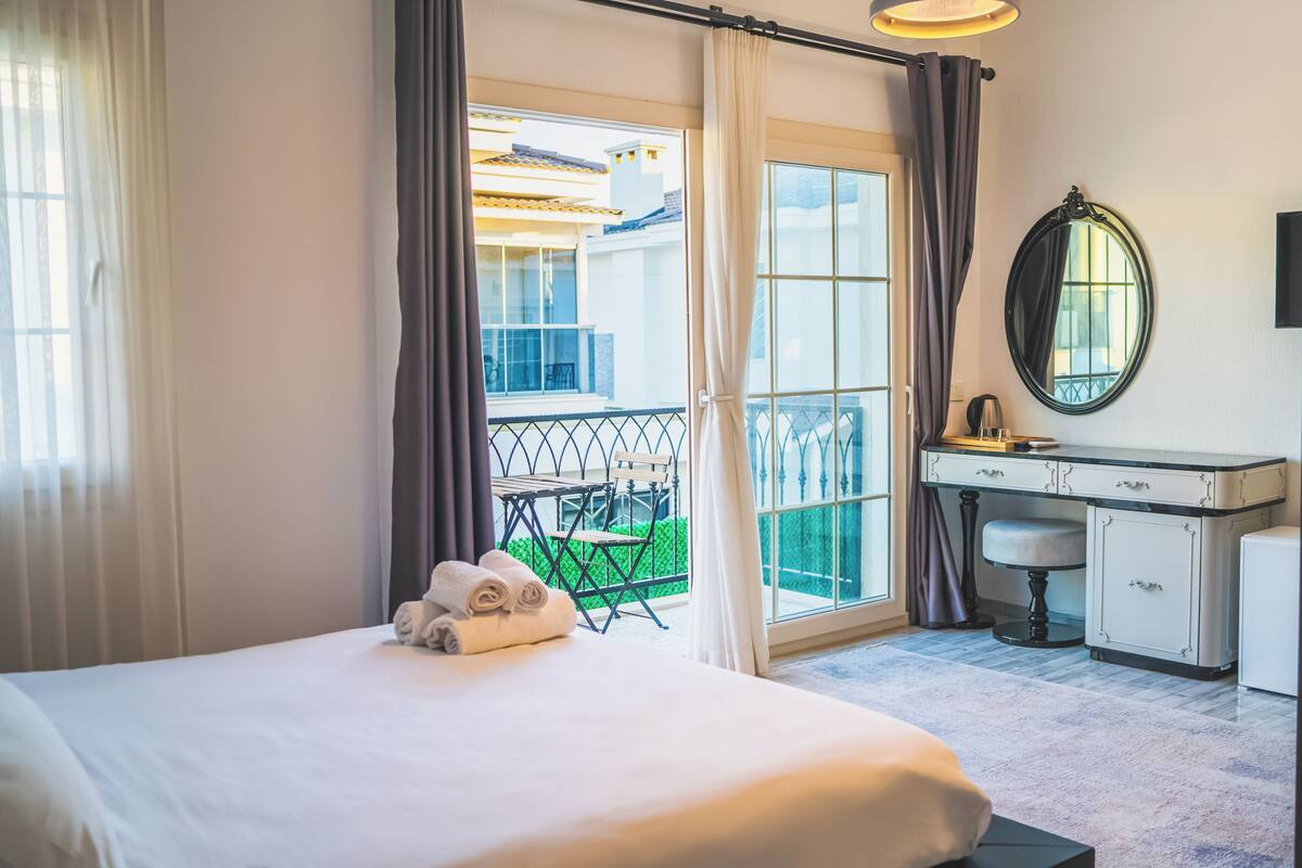 Quarto de hotel com cama, escrivaninha, cadeira, varanda com mesa e cadeiras, e uma janela grande com cortinas.