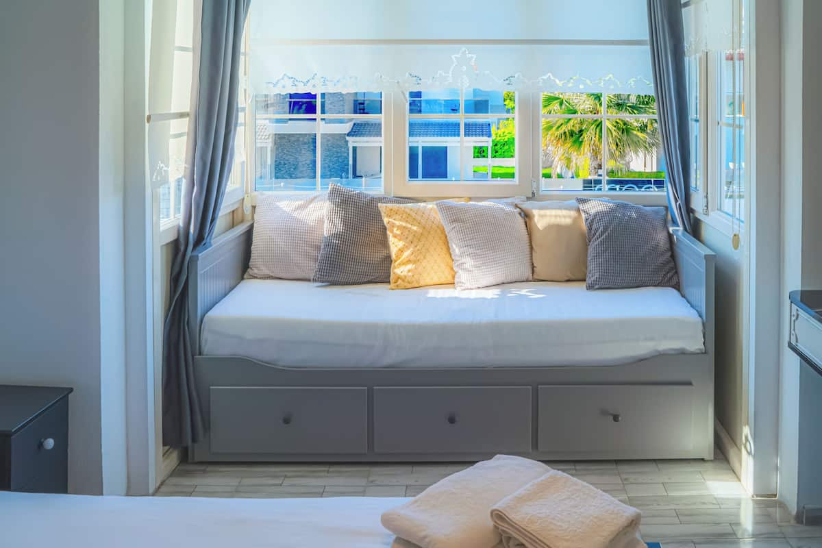 Quarto com uma janela grande, cama com várias almofadas, mesa de cabeceira e vista para edifícios e vegetação lá fora.
