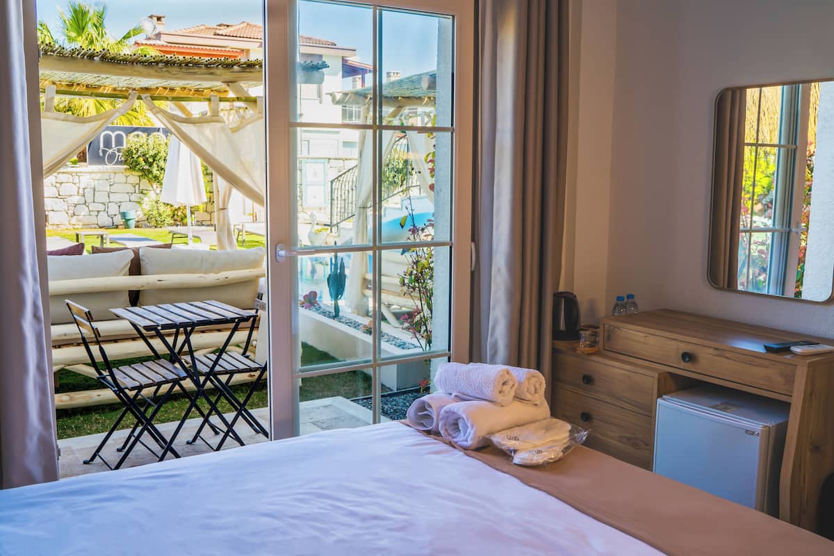 Quarto de hotel com cama, penteadeira de madeira, mini-frigorífico e janela com vista para piscina e área externa.