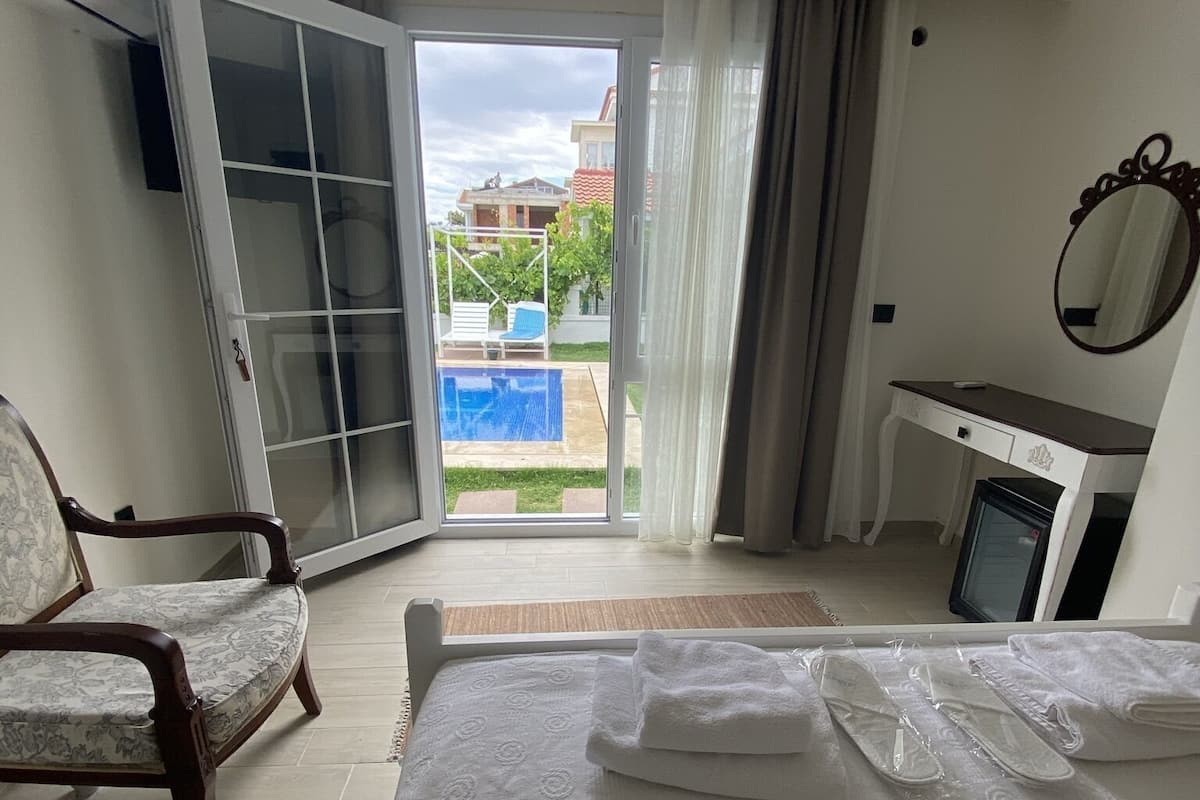 Quarto de hotel com cama, poltrona, escrivaninha, espelho e vista para uma piscina.
