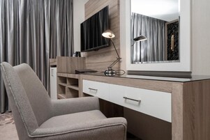 Quarto moderno com escrivaninha de madeira, cadeira cinza, espelho grande e televisão fixada na parede.