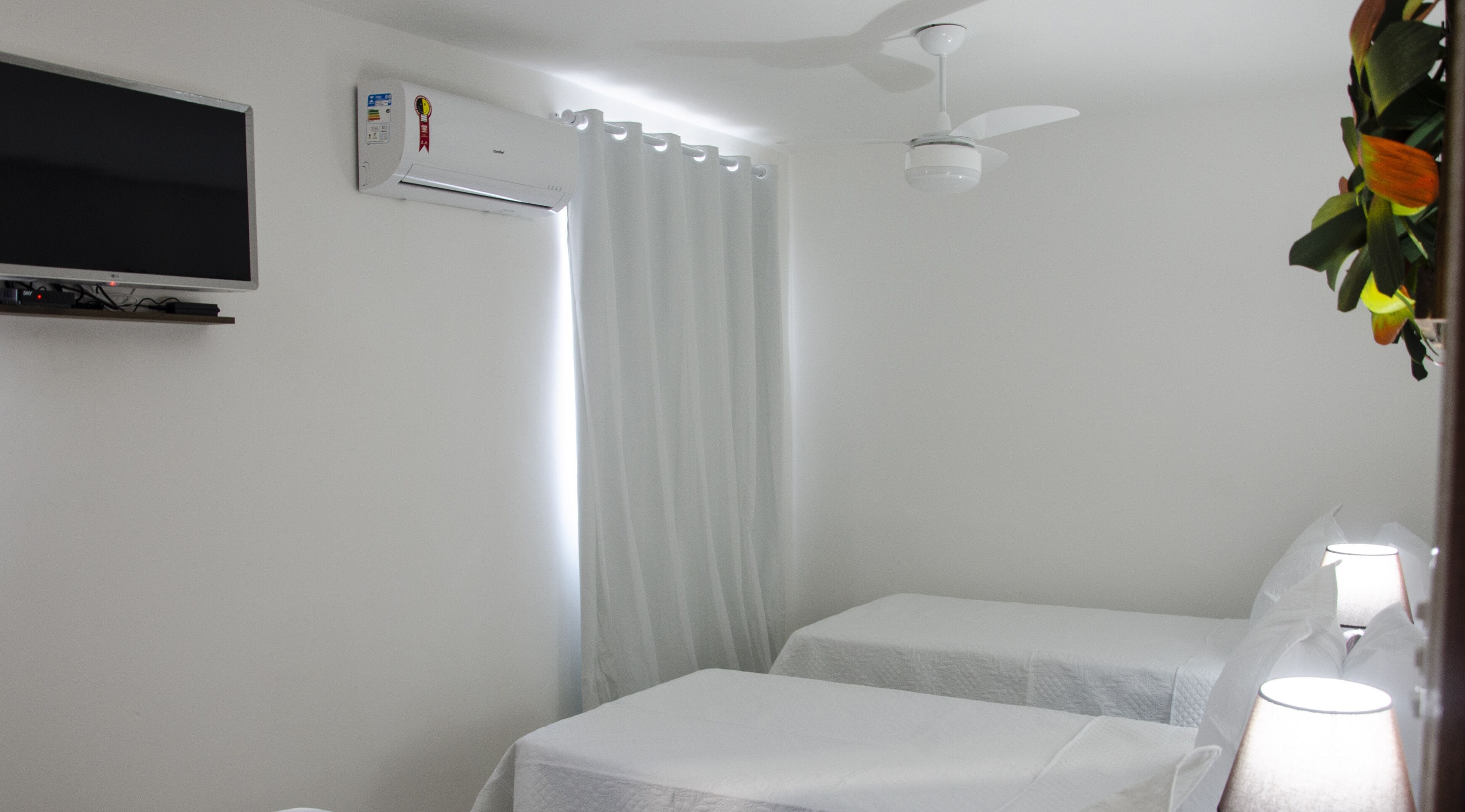 Quarto de hotel com duas camas, um ventilador de teto, ar condicionado, uma televisão e uma cortina.