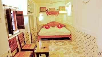 Quarto com cama, cadeira, mesinha e prateleira com objetos decorativos.
