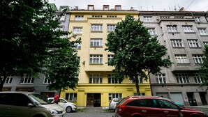 Um edifício com várias andares, fachada amarela, várias janelas e uma porta. Carros estão estacionados na frente, e árvores são visíveis em frente ao edifício.