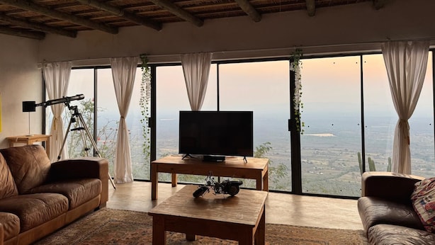 Uma sala de estar ampla com uma televisão grande sobre uma mesa de madeira, sofás de couro e um telescópio.