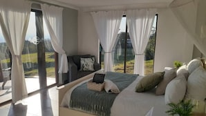 Quarto moderno com uma cama grande, um sofá e vista para uma paisagem verde através de amplas janelas.