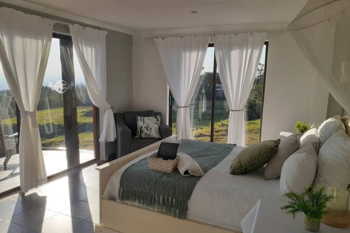 Quarto moderno com uma cama grande, um sofá e vista para uma paisagem verde através de amplas janelas.