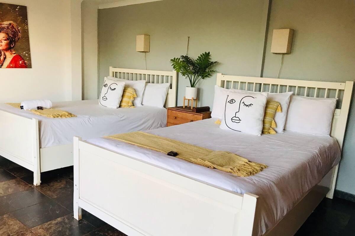 Quarto de hotel com duas camas de solteiro, uma pequena mesa de cabeceira de madeira, uma planta em vaso e um quadro na parede.