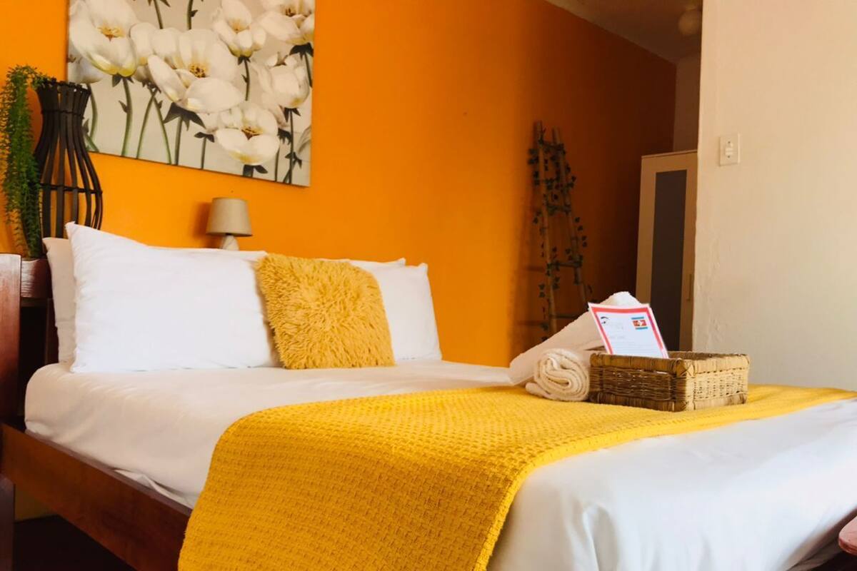 Cama bem arrumada com roupa de cama branca e amarela, uma luminária, um cesto com toalha e um quadro floral na parede.