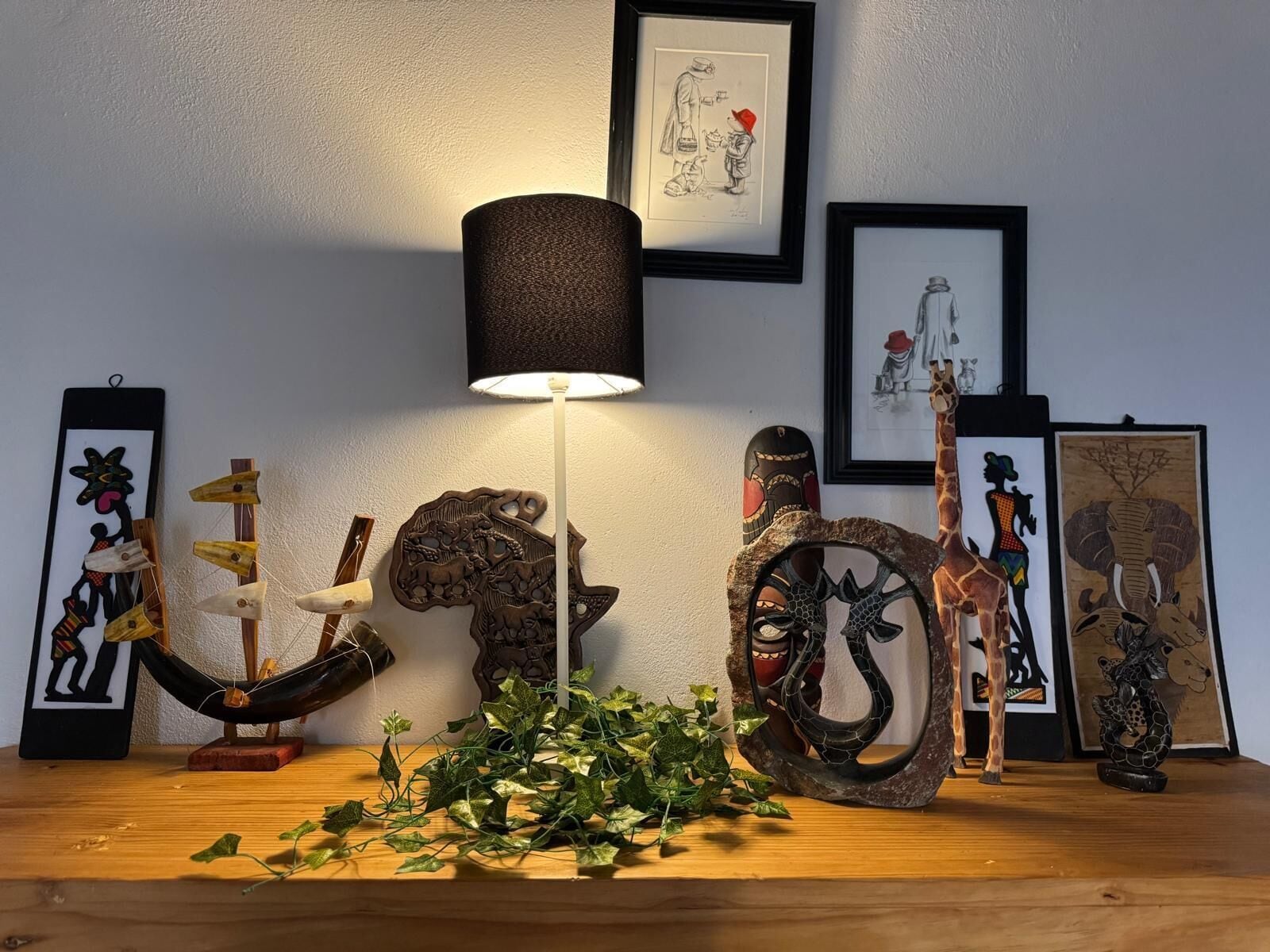 Quarto com uma mesa de madeira exibindo vários objetos decorativos, uma luminária e obras de arte emolduradas na parede.