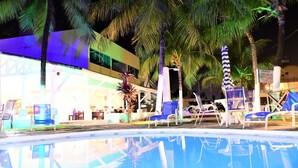 Área de piscina com espreguiçadeiras, palmeiras e um edifício com sinalização iluminada.