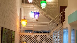 Um hall espaçoso com teto alto, paredes de tijolos brancos e uma escadaria. Há várias áreas de estar com sofás e mesas, e lustres coloridos pendem do teto.