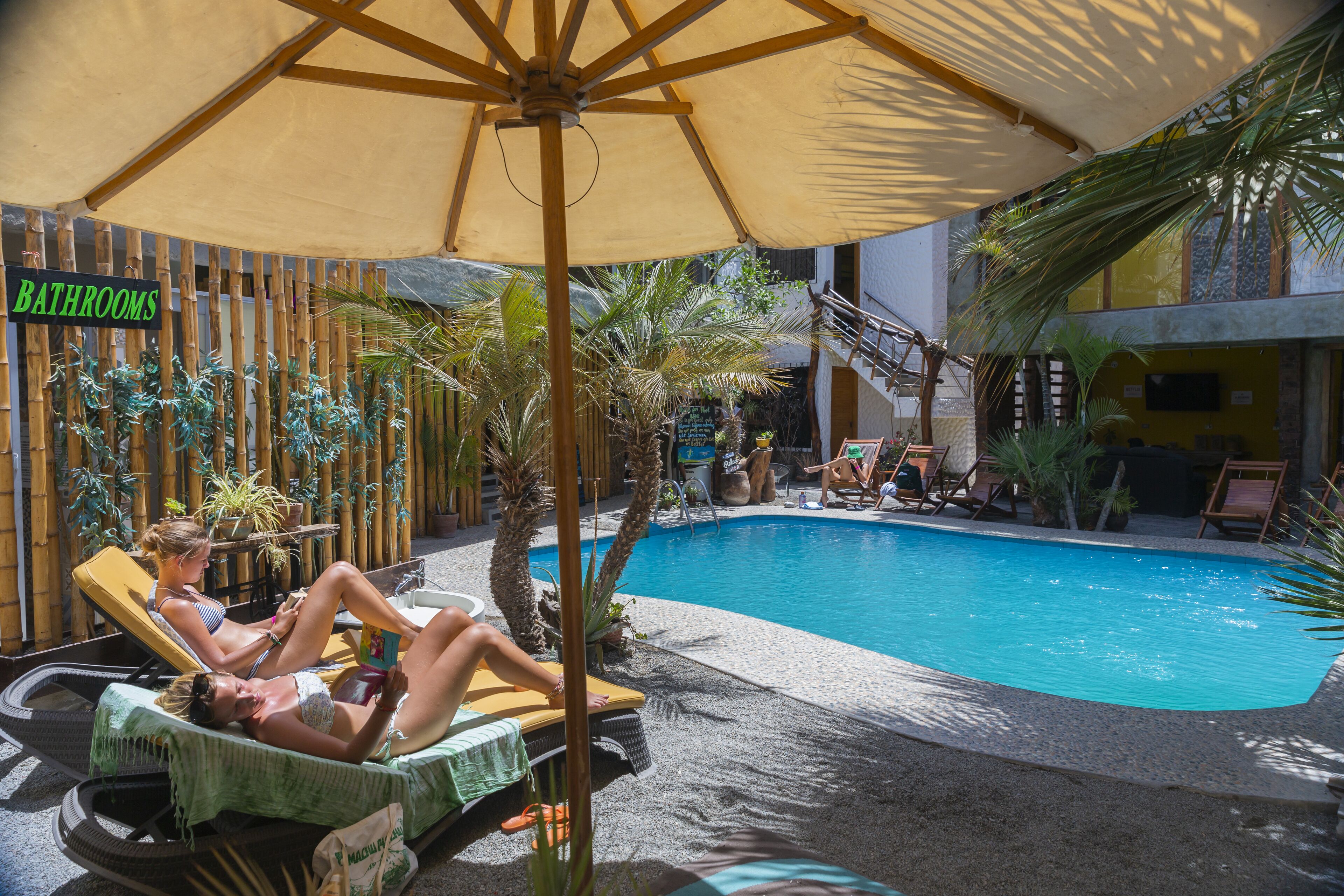 Área da piscina com espreguiçadeiras, sinalização para banheiros e plantas tropicais.