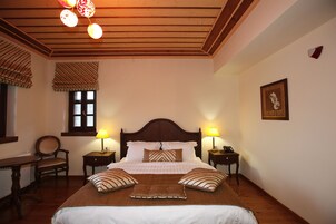 Quarto bem arrumado com teto de madeira, cama grande com almofadas listradas, duas mesas de cabeceira com luminárias, uma cadeira e uma janela com persianas.