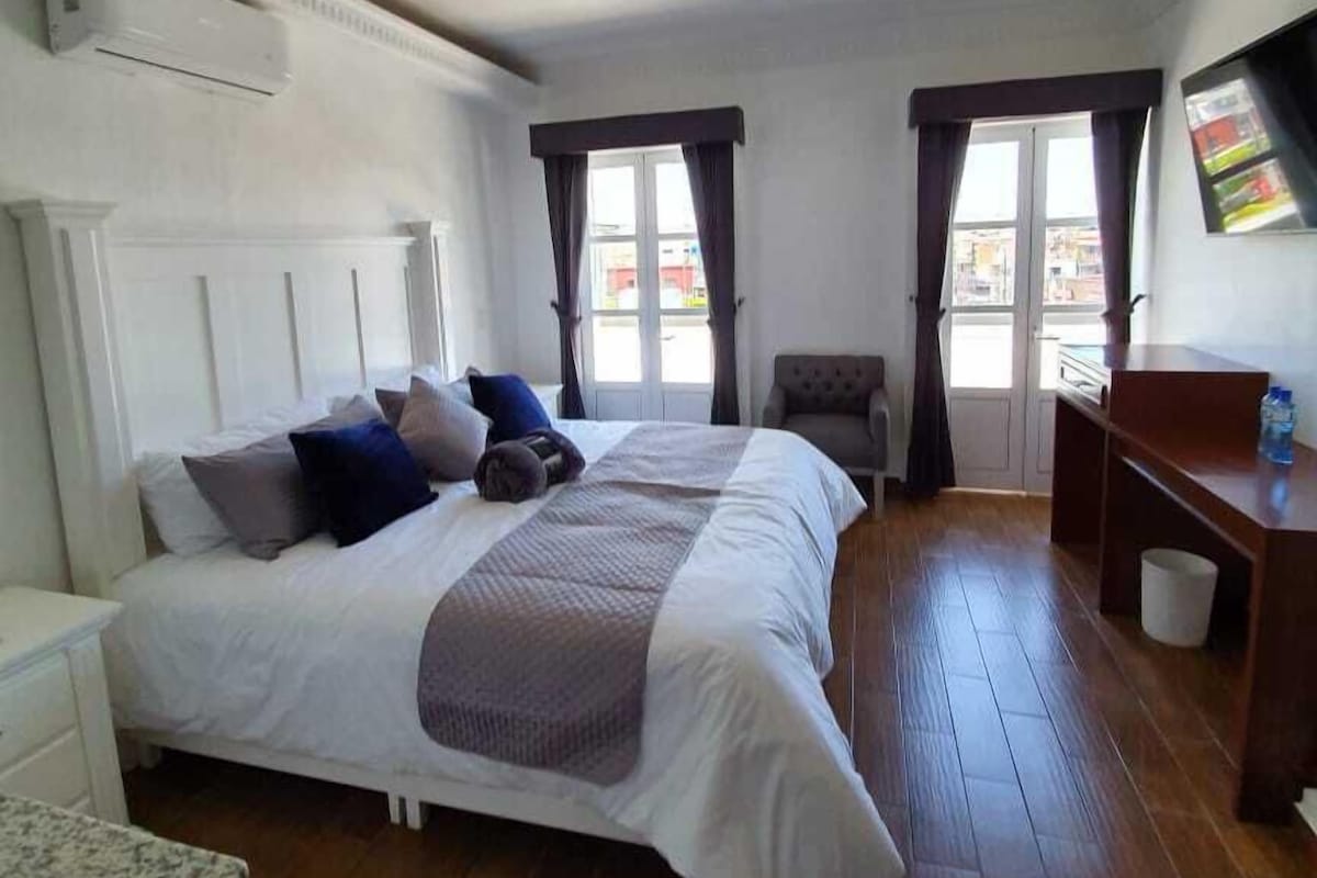 Quarto com uma cama grande, uma escrivaninha e duas janelas com cortinas.