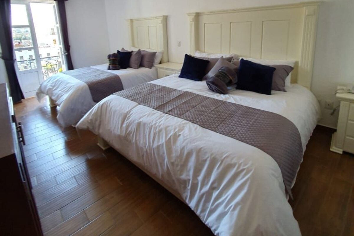 Quarto de hotel com duas camas, piso de madeira e janela com cortinas.