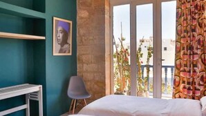 Quarto com cama, escrivaninha, cadeira e janela com vista para edifícios e plantas.