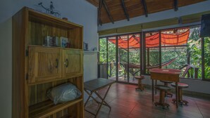 Quarto com estante de madeira, mesa de jantar com bancos e vista para vegetação exuberante através de grandes janelas.