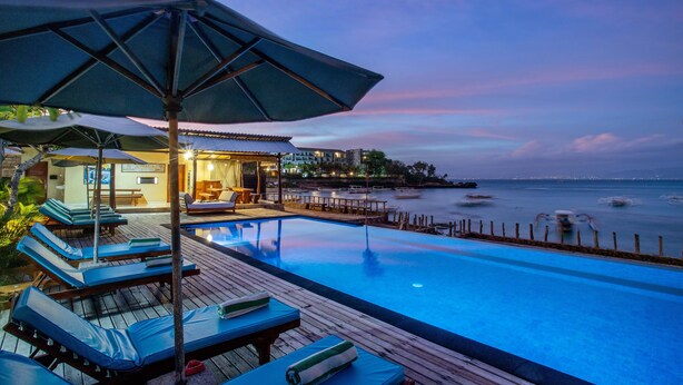 Área ao lado da piscina com espreguiçadeiras e um guarda-sol, com vista para um corpo de água ao entardecer.
