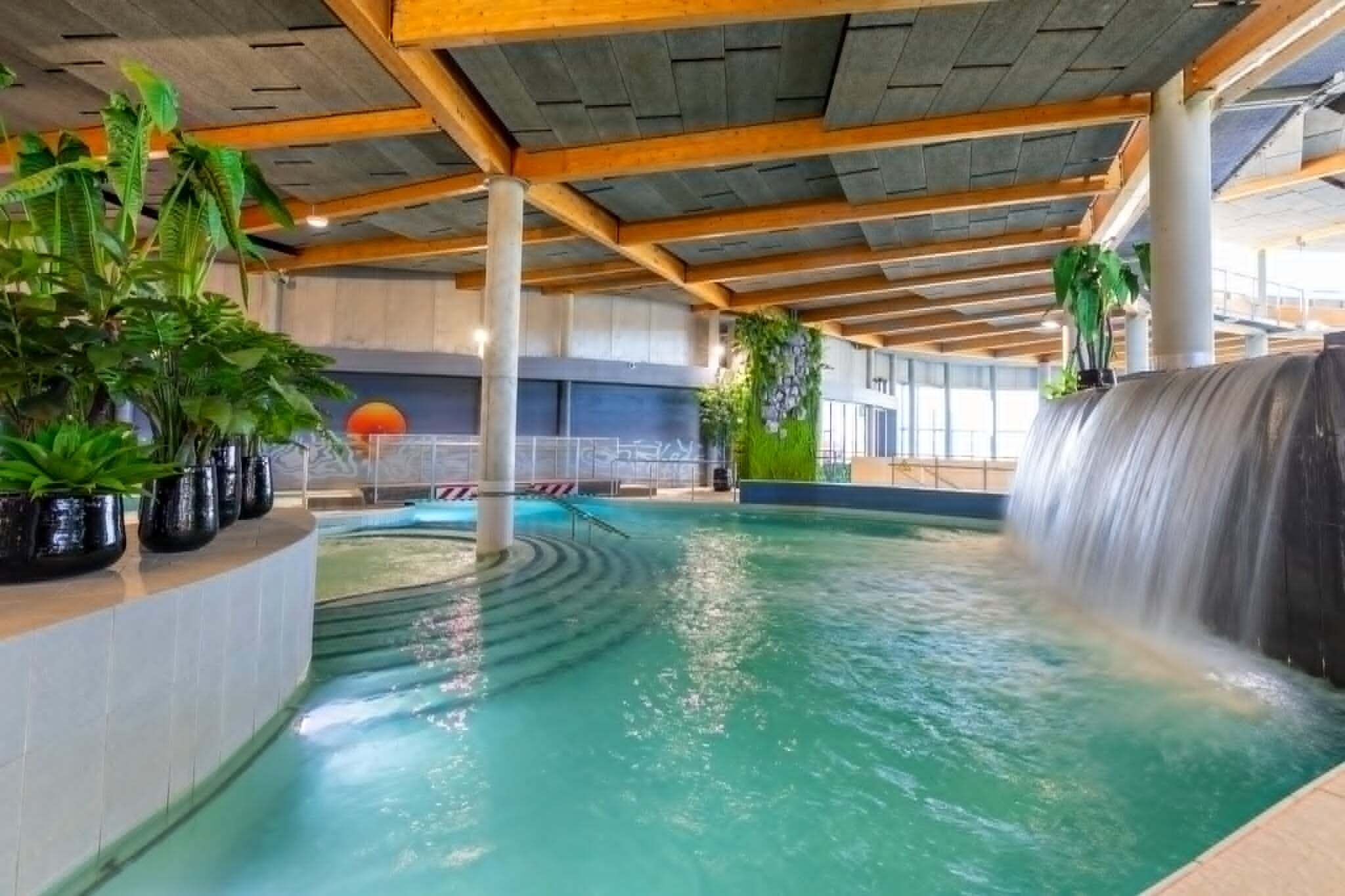 Piscina interna com cachoeira, cercada por plantas em vasos e teto alto com vigas de madeira.
