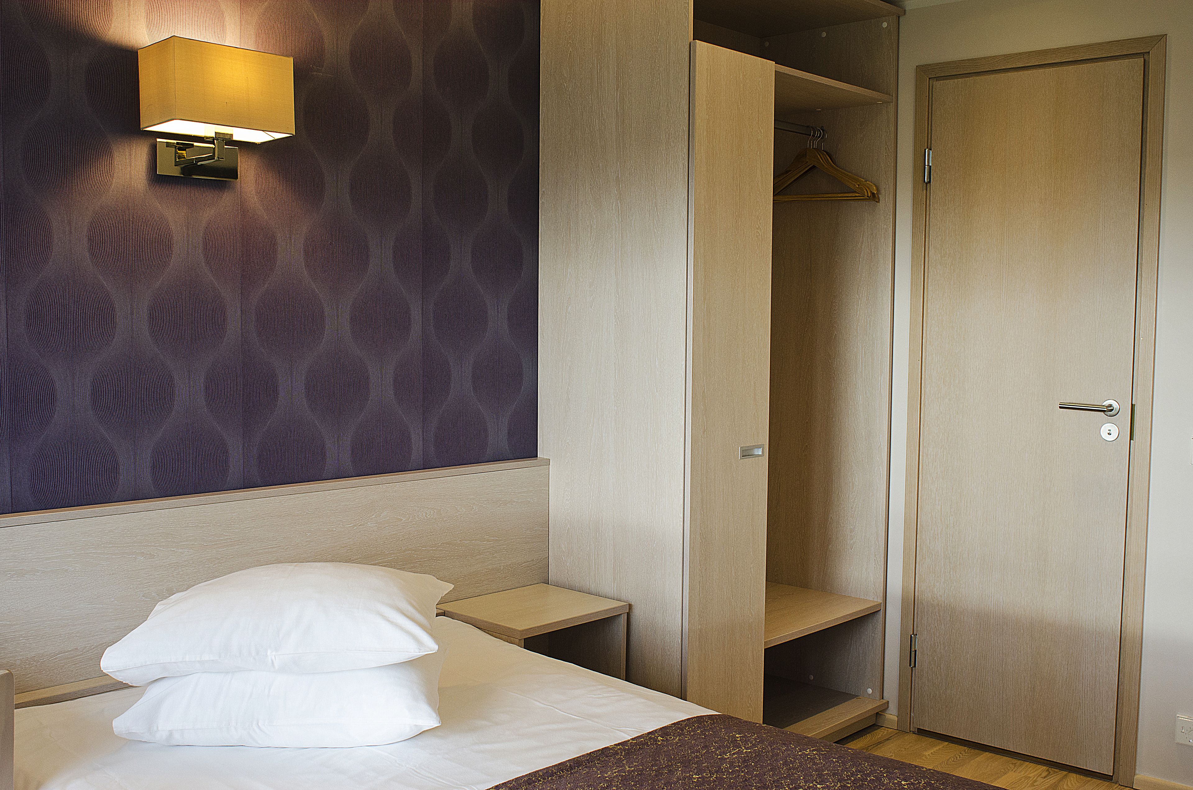 Quarto de hotel com cama, mesa de cabeceira, guarda-roupa e luminária fixada na parede.