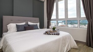 Cama bem arrumada com edredom branco e travesseiros, cabeceira cinza e vista para a cidade através de janelas amplas com cortinas.