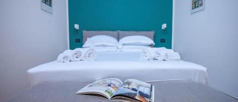 Uma cama bem arrumada com roupa de cama e almofadas brancas, e uma revista sobre a cama, com paredes cor de verde-água.
