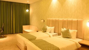 Quarto de hotel com duas camas, cortina verde, chão de madeira e mesa de cabeceira com abajur.