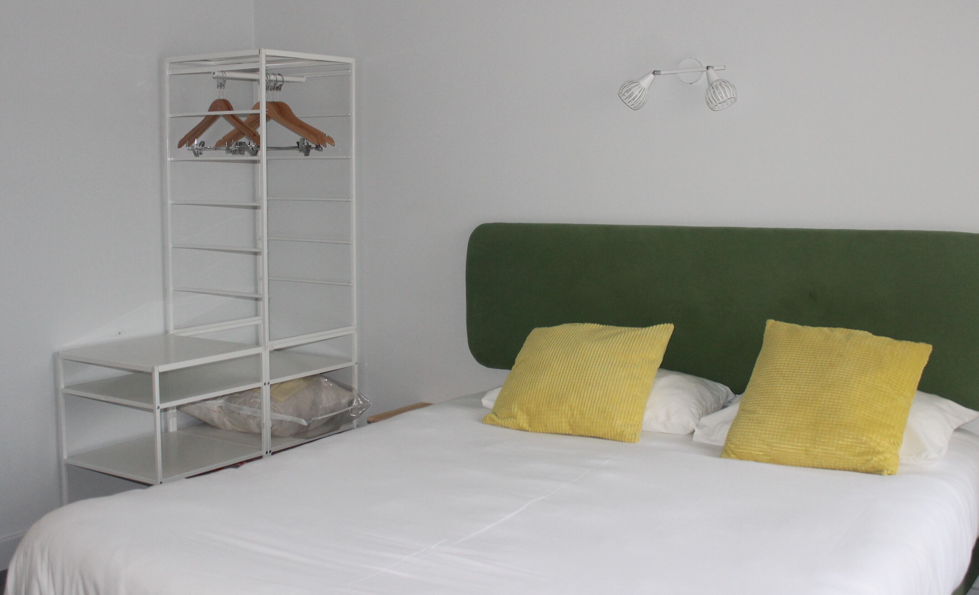 Quarto com cama, dois travesseiros amarelos, cabeceira verde, guarda-roupa branco e luminária fixada na parede.