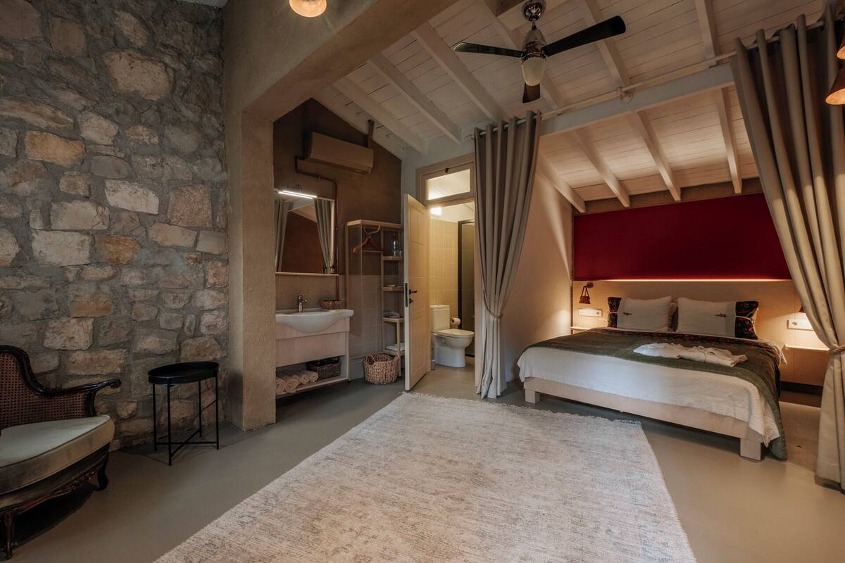 Um quarto com parede de pedra, teto de madeira, cama com cabeceira vermelha, mesa de cabeceira e luminária pendente.