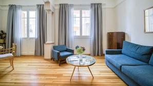 Uma sala de estar com piso de madeira, um sofá azul, uma poltrona azul, uma mesa de centro redonda com um vaso de flores e um armário de madeira.