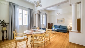 Uma sala de estar ampla com uma mesa de jantar redonda de madeira, dois sofás azuis e uma janela grande com cortinas cinzentas.