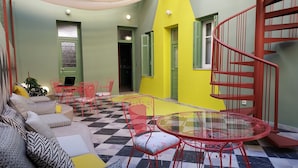 Um quarto com piso xadrez, cadeiras de metal vermelhas, uma mesa de vidro e um sofá com almofadas estampadas.