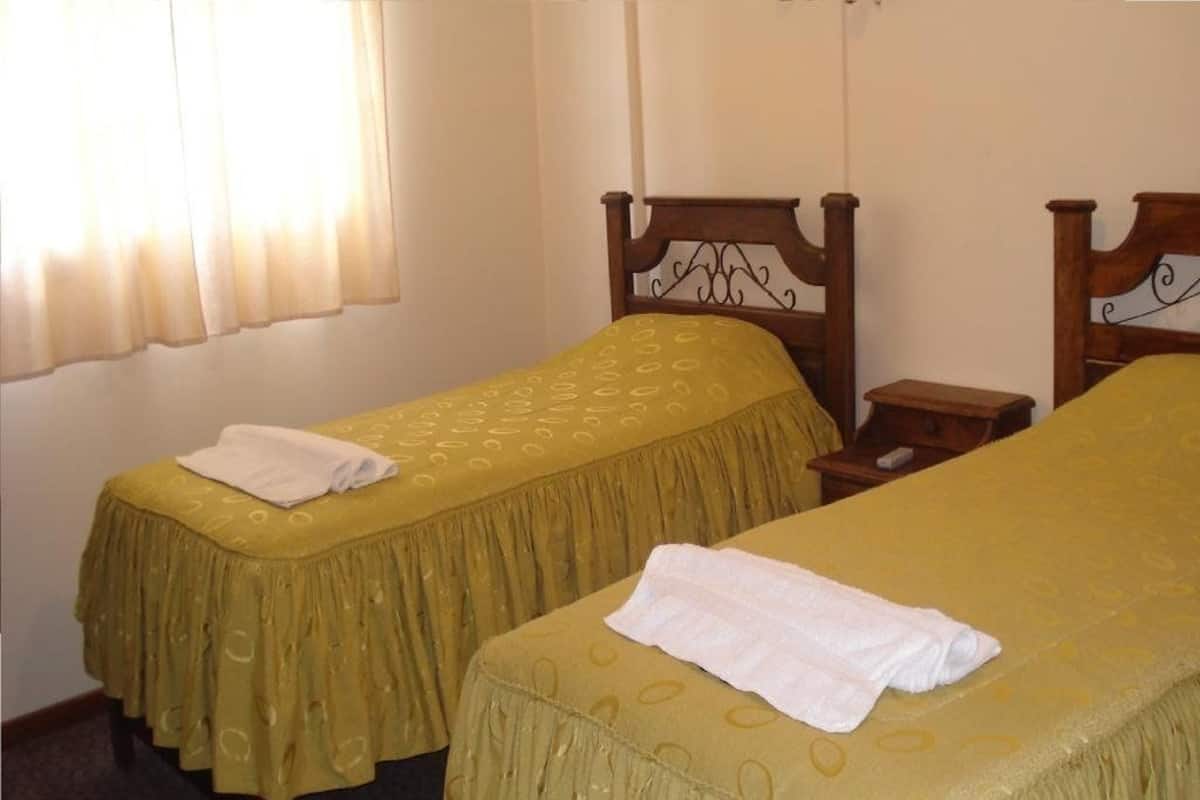 Duas camas de solteiro com cabeceiras de madeira, uma mesinha de cabeceira e uma janela com cortinas.
