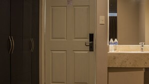 Quarto de hotel com porta fechada, banheiro com pia e espelho, e piso revestido com azulejos.