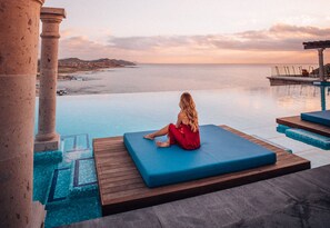 Uma pessoa sentada em um travesseiro azul perto de uma piscina, com vista para o mar e colinas ao longe.
