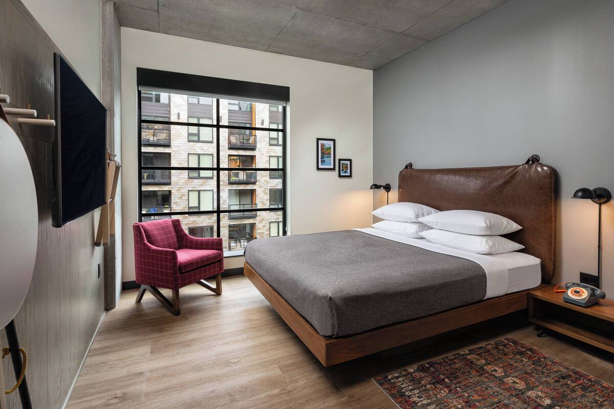 Quarto com cama, cadeira, mesa de cabeceira, luminária e janela com vista para edifícios.