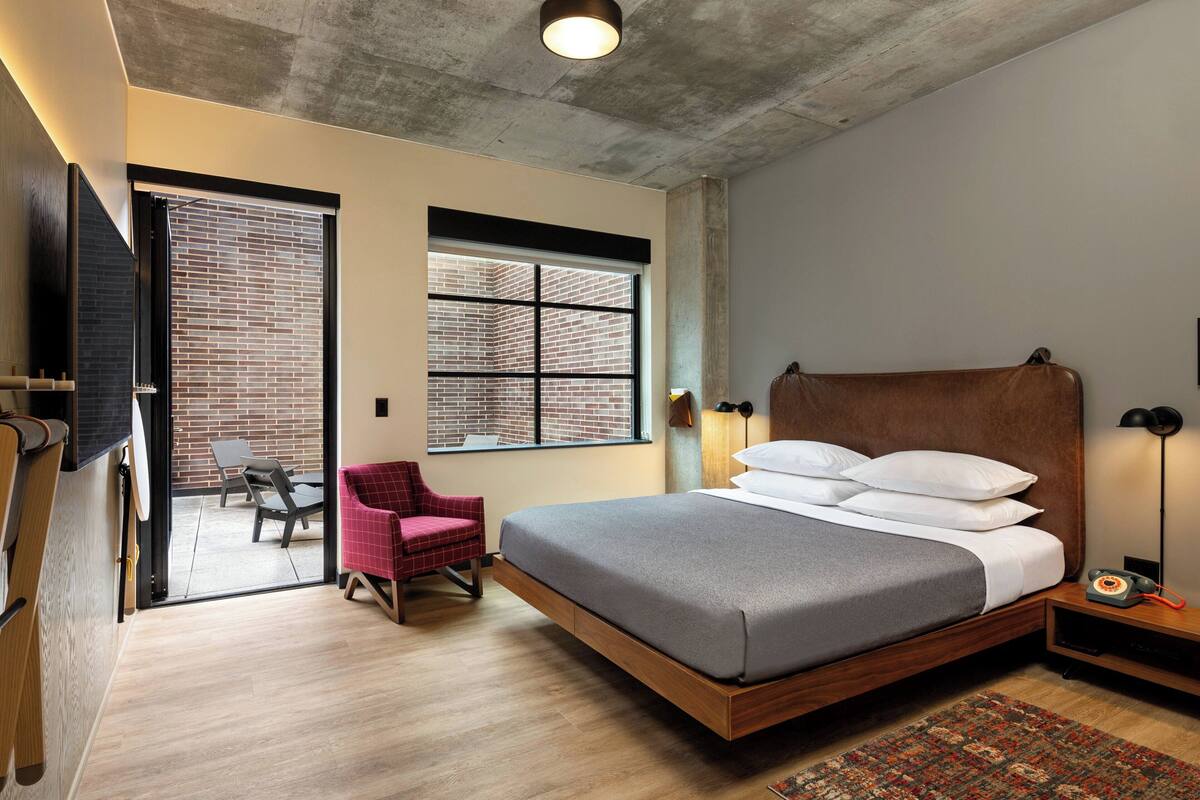 Quarto moderno com uma cama grande, uma cadeira vermelha, uma escrivaninha e uma janela com vista para uma parede de tijolos.