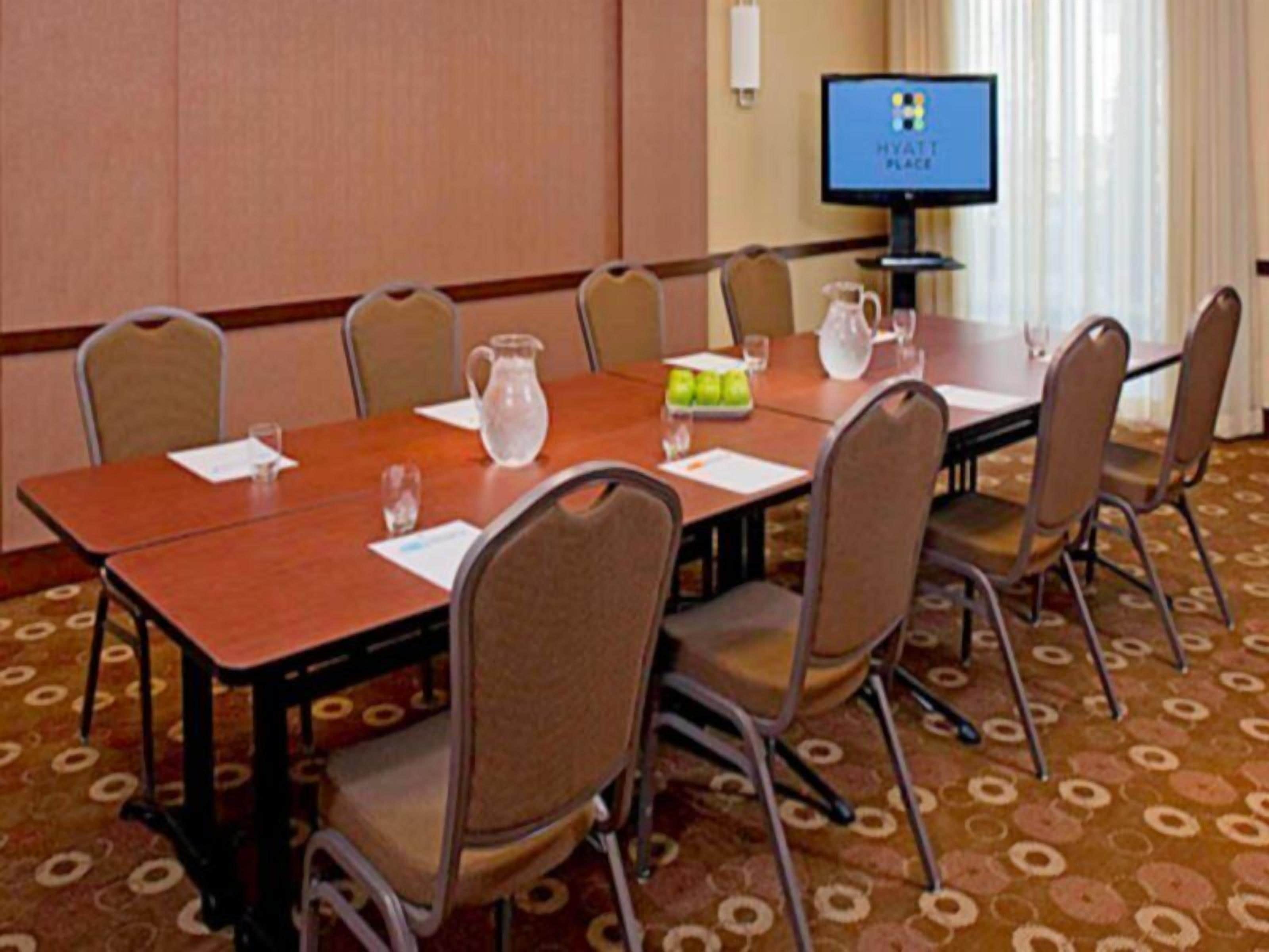 Uma sala de conferências com uma mesa comprida, seis cadeiras, um jarro, copos e um monitor exibindo 'HYATT PLACE'.