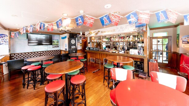 Uma área de bar com mesas e cadeiras de madeira, balcão com diversos frascos e uma tela de televisão.