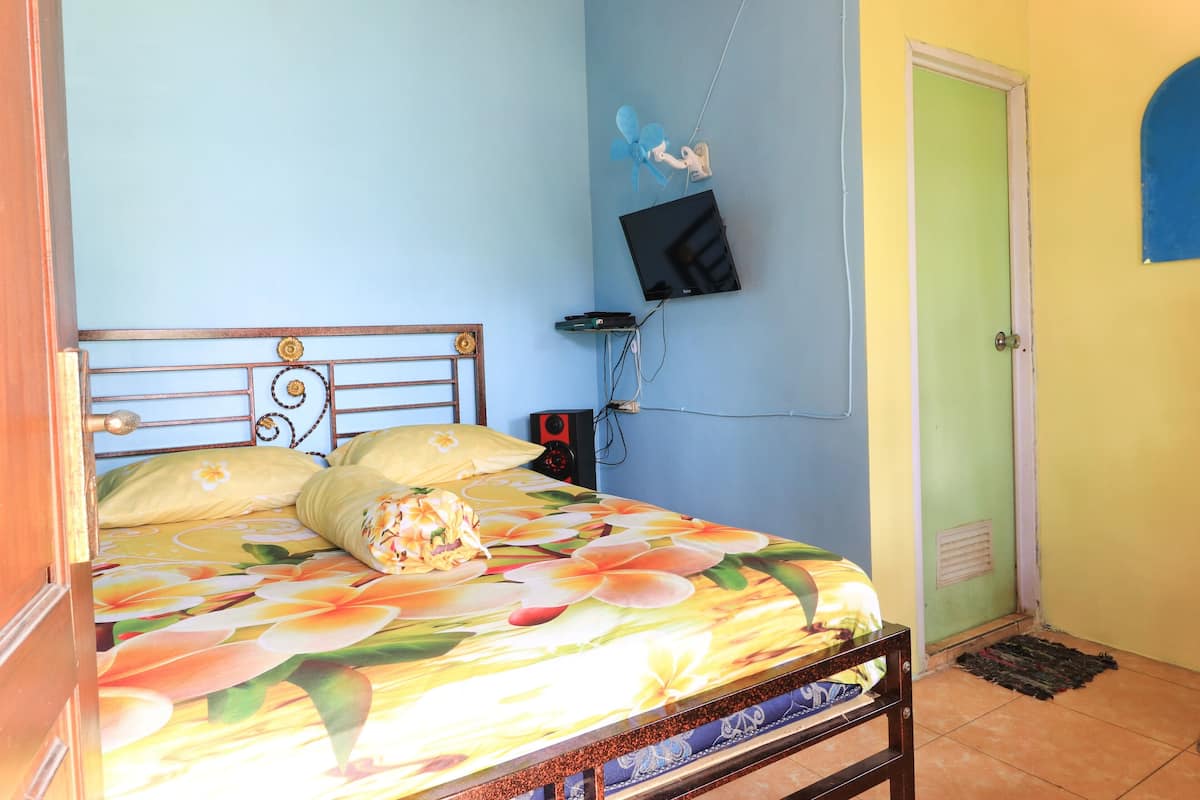 Uma cama com colcha florida, uma televisão fixada na parede e uma porta com janela.
