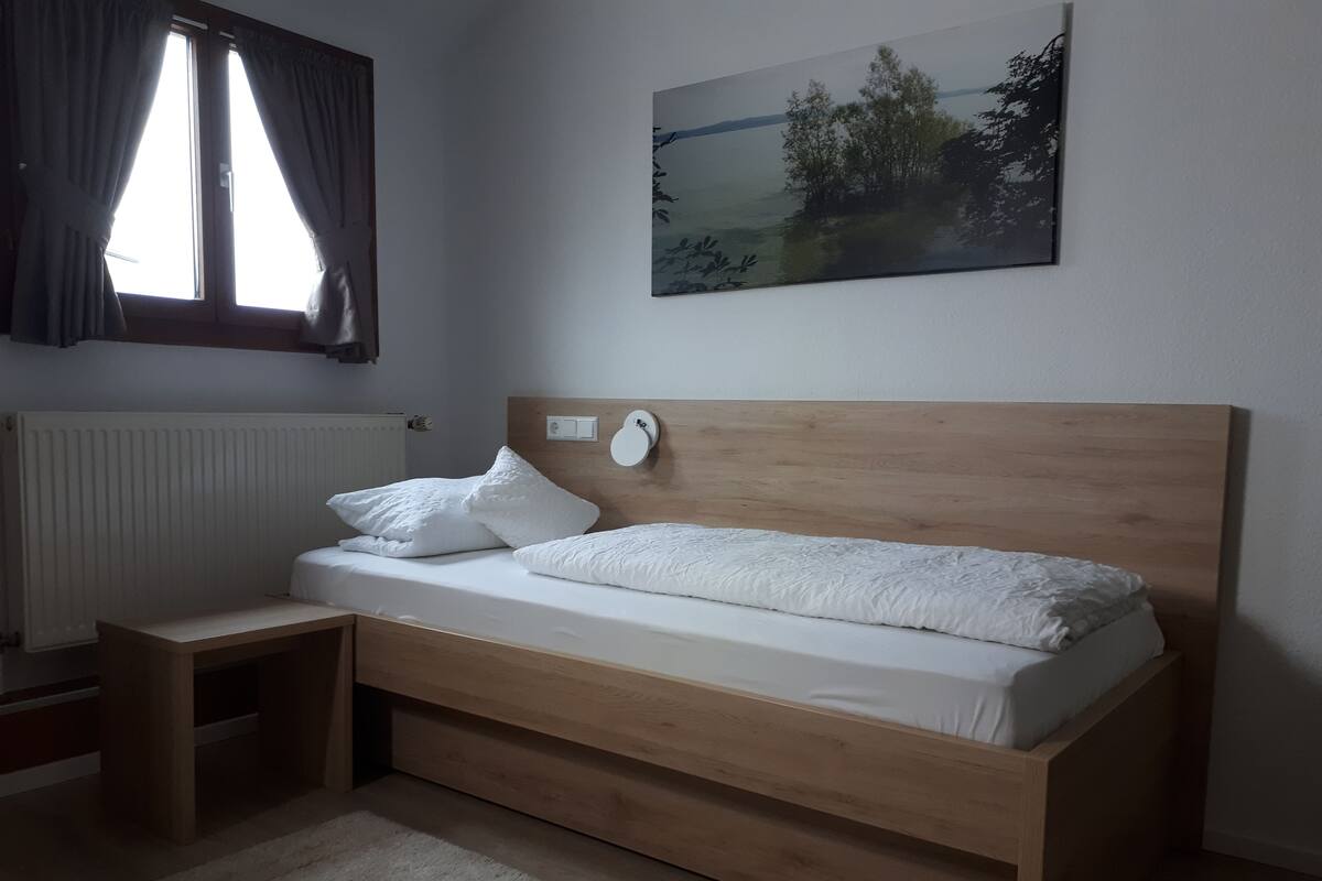 Quarto individual com cama de madeira, roupa de cama branca, mesa de cabeceira e janela com cortinas.
