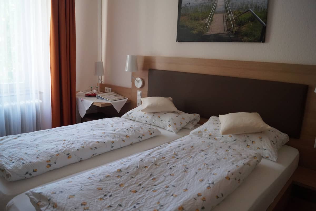 Quarto de hotel com duas camas, uma mesinha de cabeceira com abajur, um quadro na parede e uma janela com cortinas.