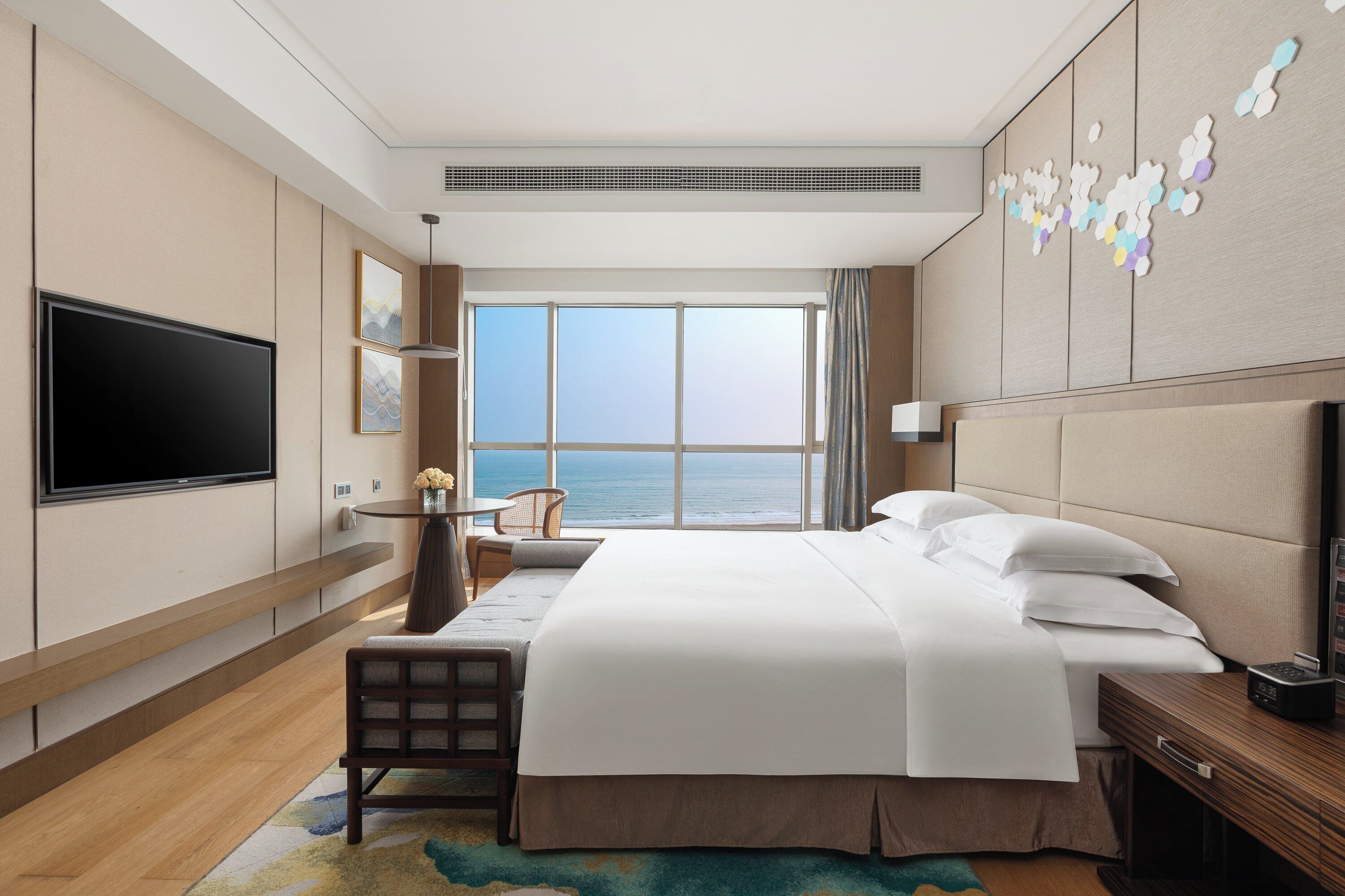 Quarto de hotel com uma cama grande, televisão, escrivaninha com cadeira, vista para o mar e uma peça de arte decorativa na parede.