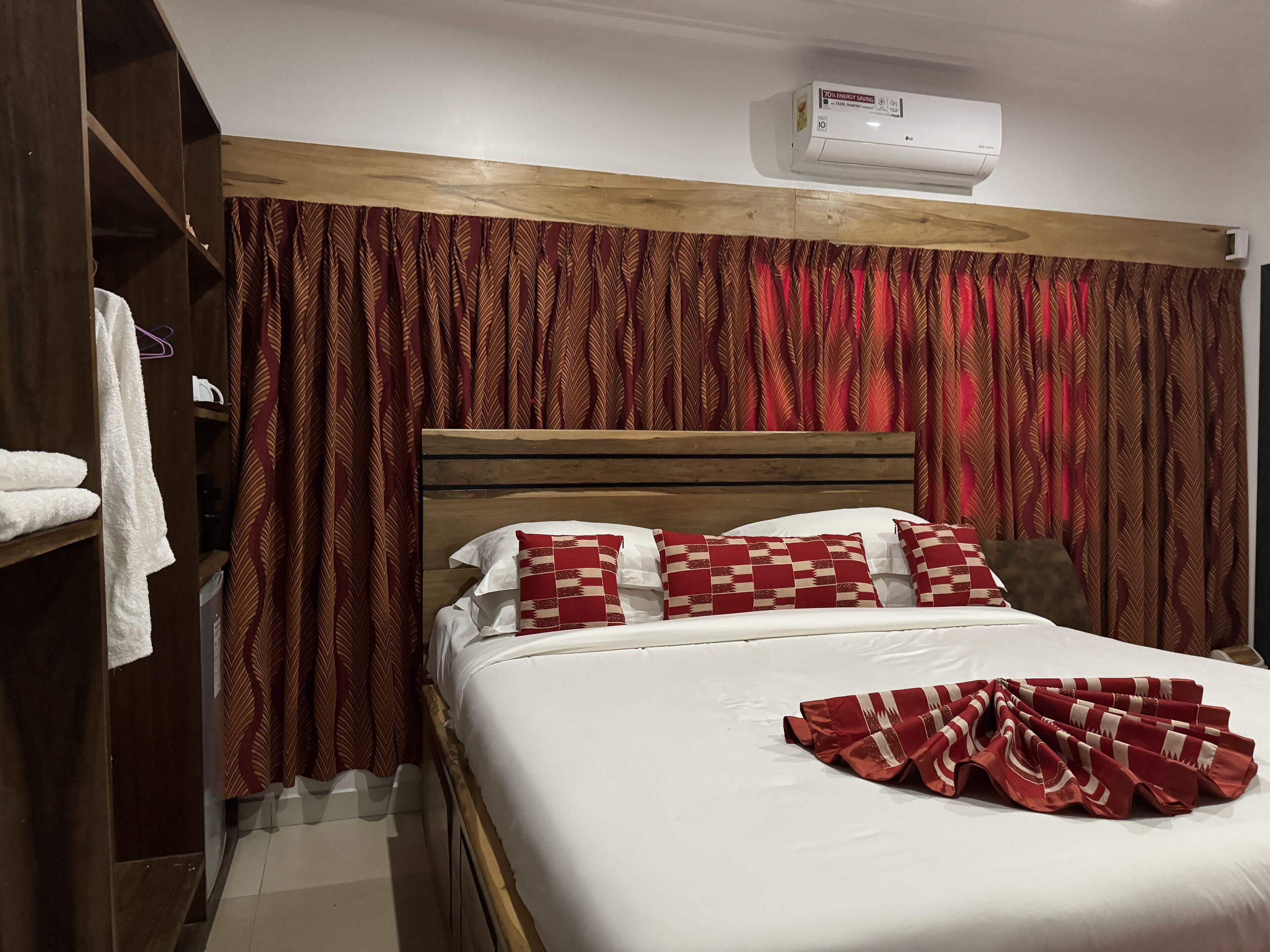 Quarto de hotel com cabeceira de madeira, cama com roupa de cama branca e almofadas estampadas vermelhas, cortina vermelha e dourada e um ar condicionado na parede.